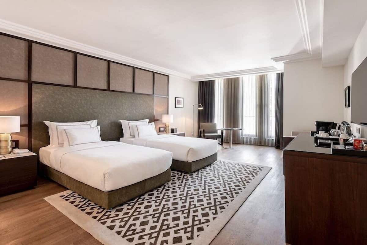 Quarto de hotel com duas camas, uma escrivaninha, uma televisão e uma janela grande.