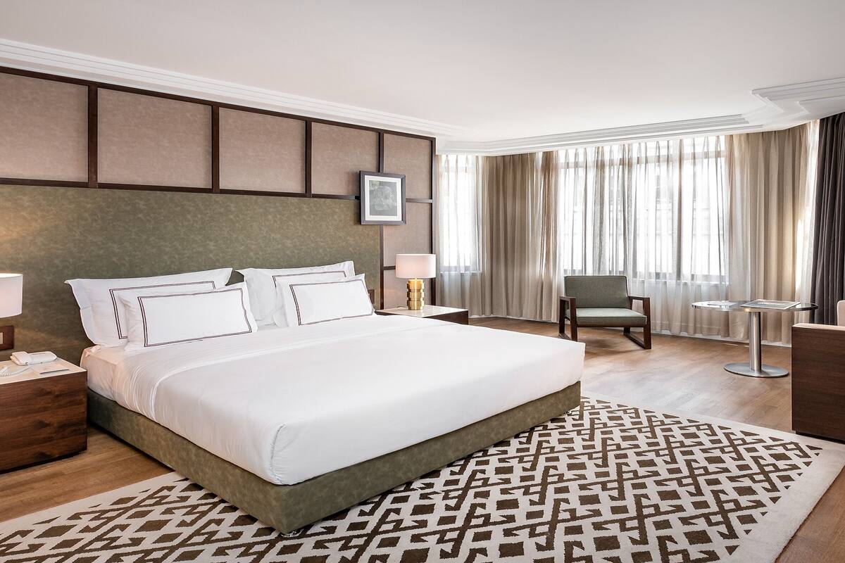 Quarto de hotel com uma cama grande, mesas de cabeceira, uma cadeira, uma pequena mesa e uma janela com cortinas.