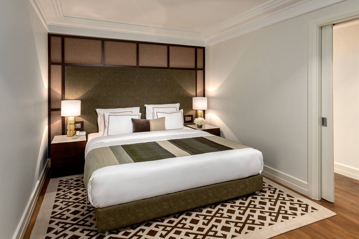 Quarto de hotel com uma cama grande, duas mesas de cabeceira com abajures, um painel de cabeceira decorativo e um tapete estampado.