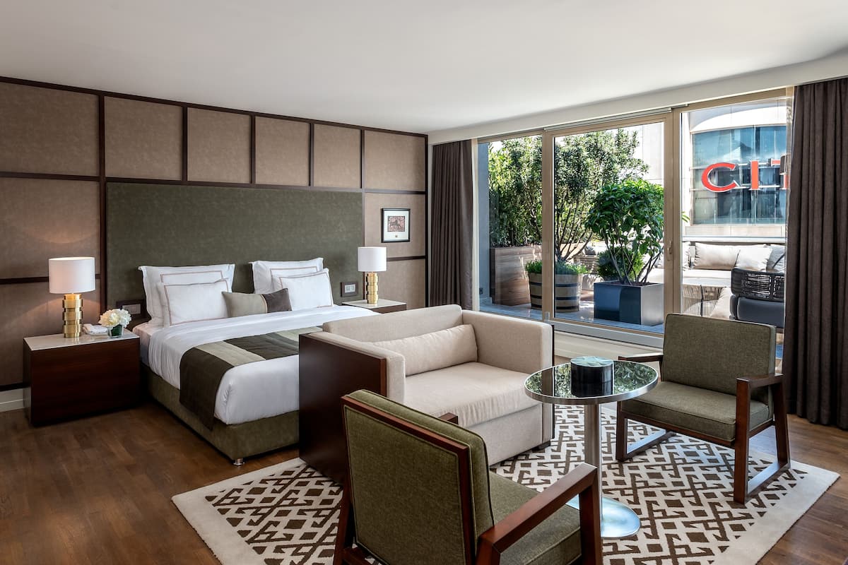 Quarto de hotel com uma cama, duas cadeiras, uma mesinha e uma mesa de cabeceira com abajur.