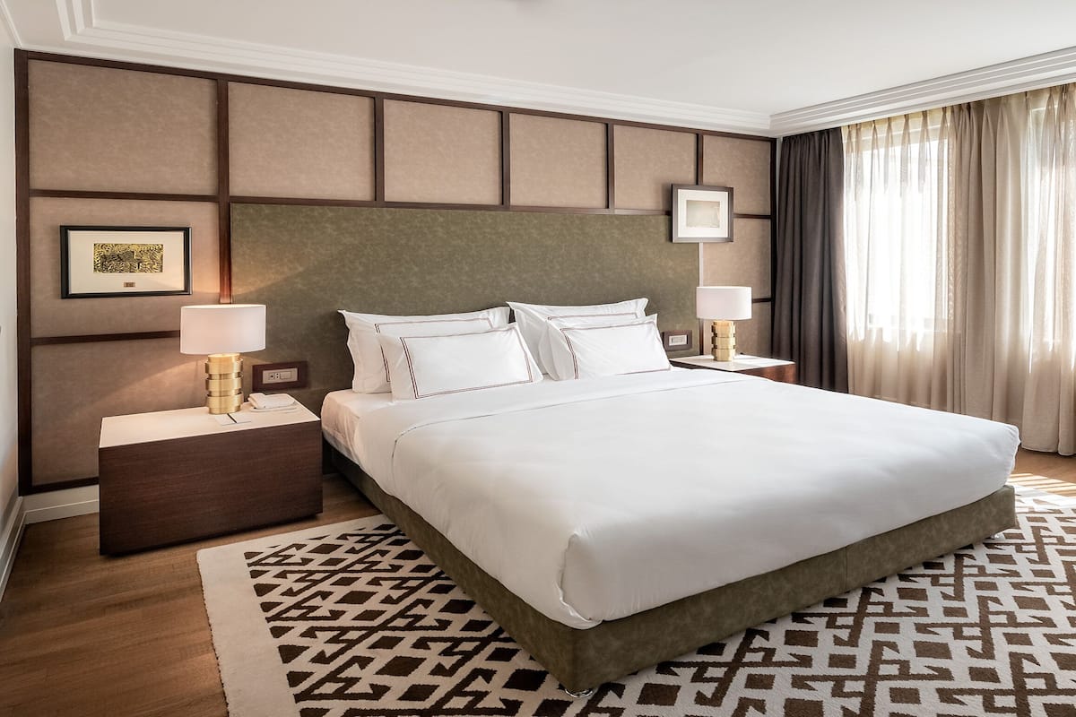 Quarto de hotel com uma cama grande, duas mesas de cabeceira com abajures, um tapete estampado e vista para o exterior através de cortinas finas.