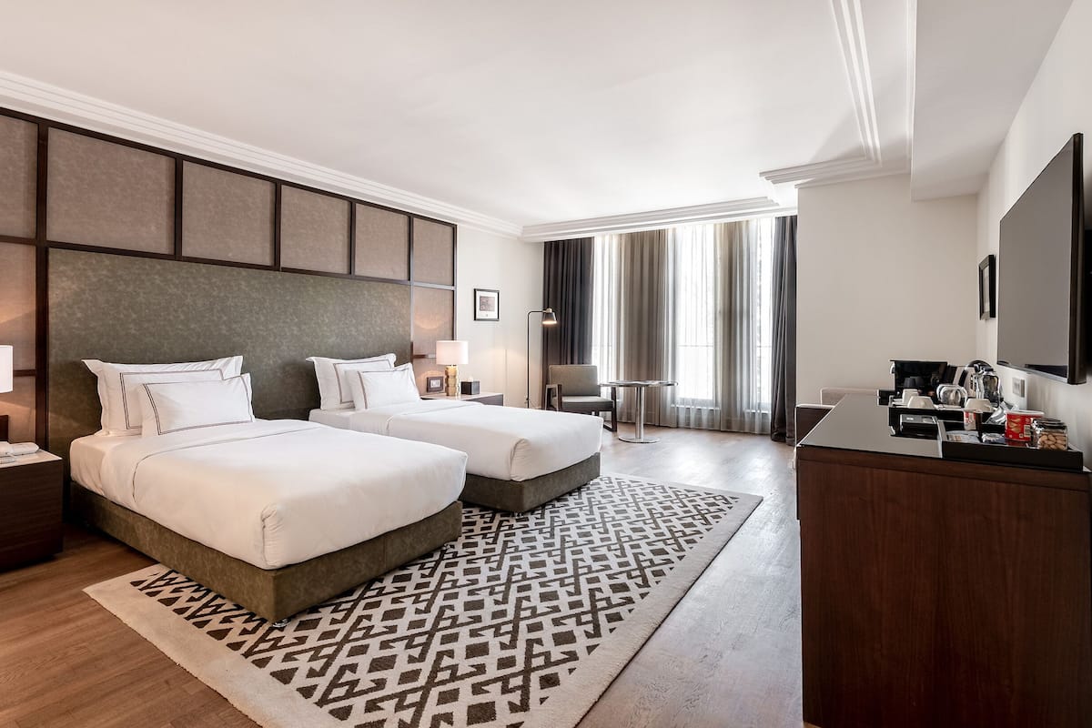 Quarto de hotel com duas camas, uma escrivaninha, uma cadeira, uma televisão e uma janela grande com cortinas.