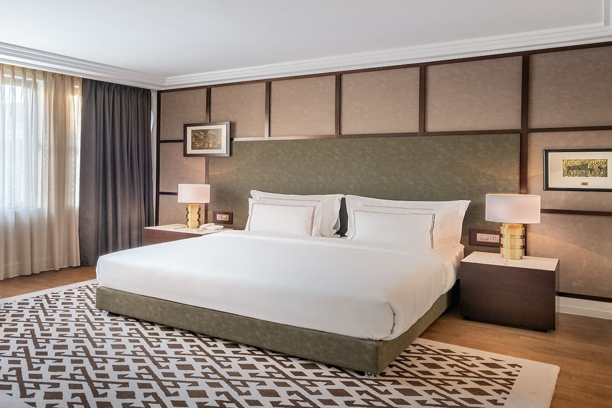 Quarto de hotel com uma cama grande, duas mesas de cabeceira com luminárias, uma poltrona e um tapete estampado.