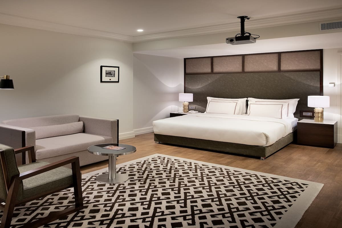 Quarto de hotel moderno com uma cama grande, um sofá, uma mesinha e um tapete estampado.