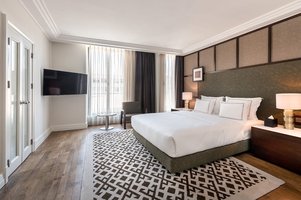 Quarto de hotel moderno com uma cama grande, uma TV de tela plana, uma mesa de cabeceira com abajur e um tapete estampado.