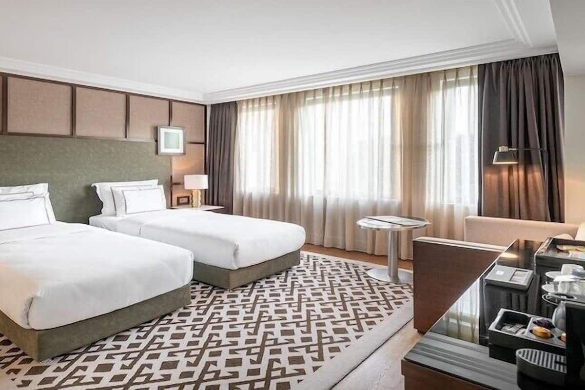 Quarto de hotel com duas camas, uma escrivaninha, uma cadeira, uma mesinha e uma televisão.