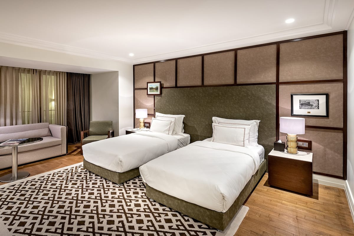 Quarto de hotel com duas camas, um sofá, uma escrivaninha, uma luminária e um tapete estampado.
