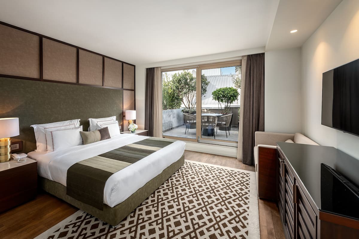 Quarto de hotel com uma cama grande, televisão, secretária e varanda com mobiliário exterior.