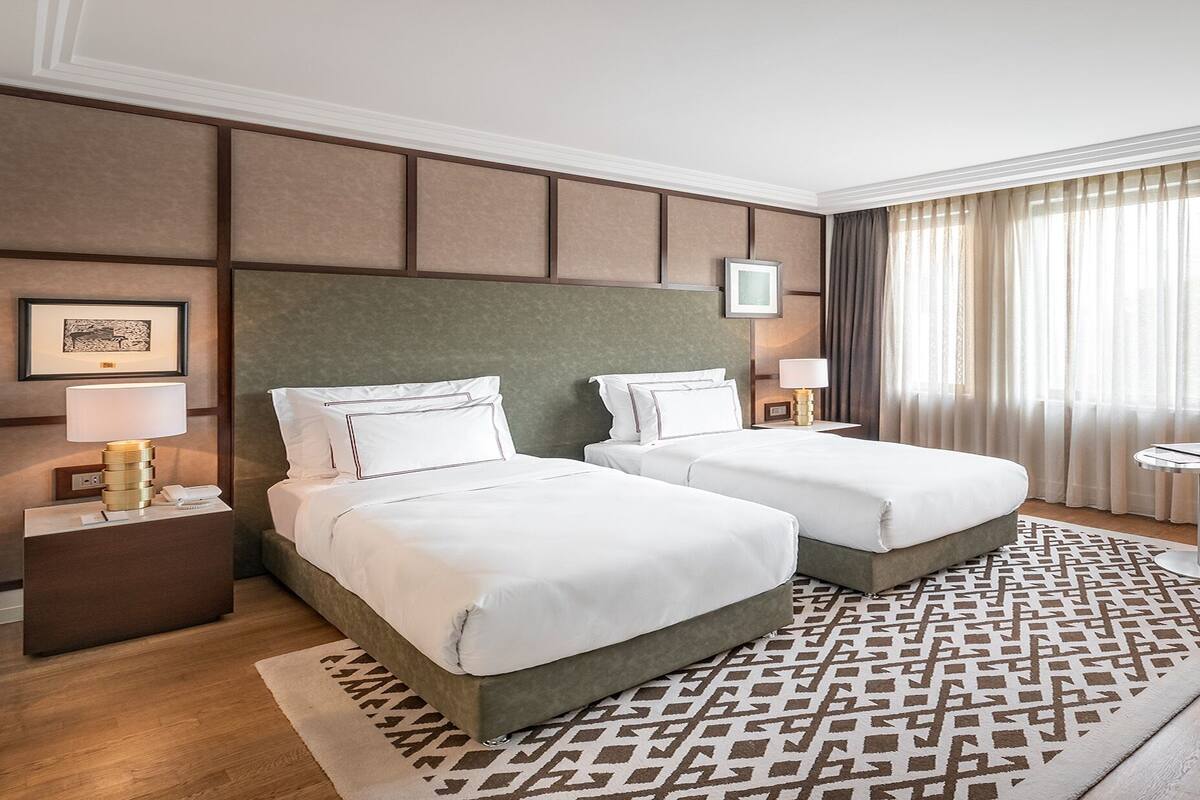 Quarto de hotel com duas camas, uma escrivaninha com candeeiro, uma mesinha e vista da cidade por grandes janelas.