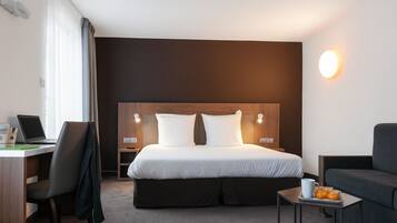 Quarto de hotel com uma cama grande, uma escrivaninha com um laptop, uma cadeira, uma pequena mesa com uma xícara e laranjas, e um criado-mudo com uma luminária.