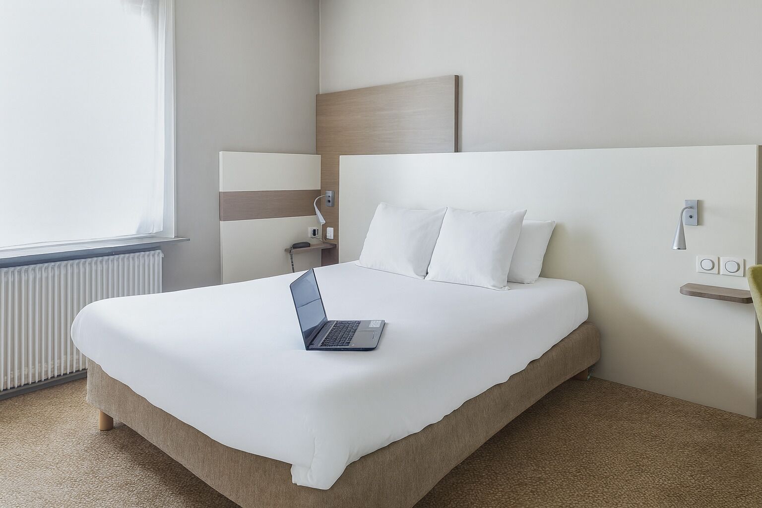 Quarto de hotel com uma cama grande, um laptop sobre a cama, uma luminária de cabeceira e uma janela com vista para o céu.
