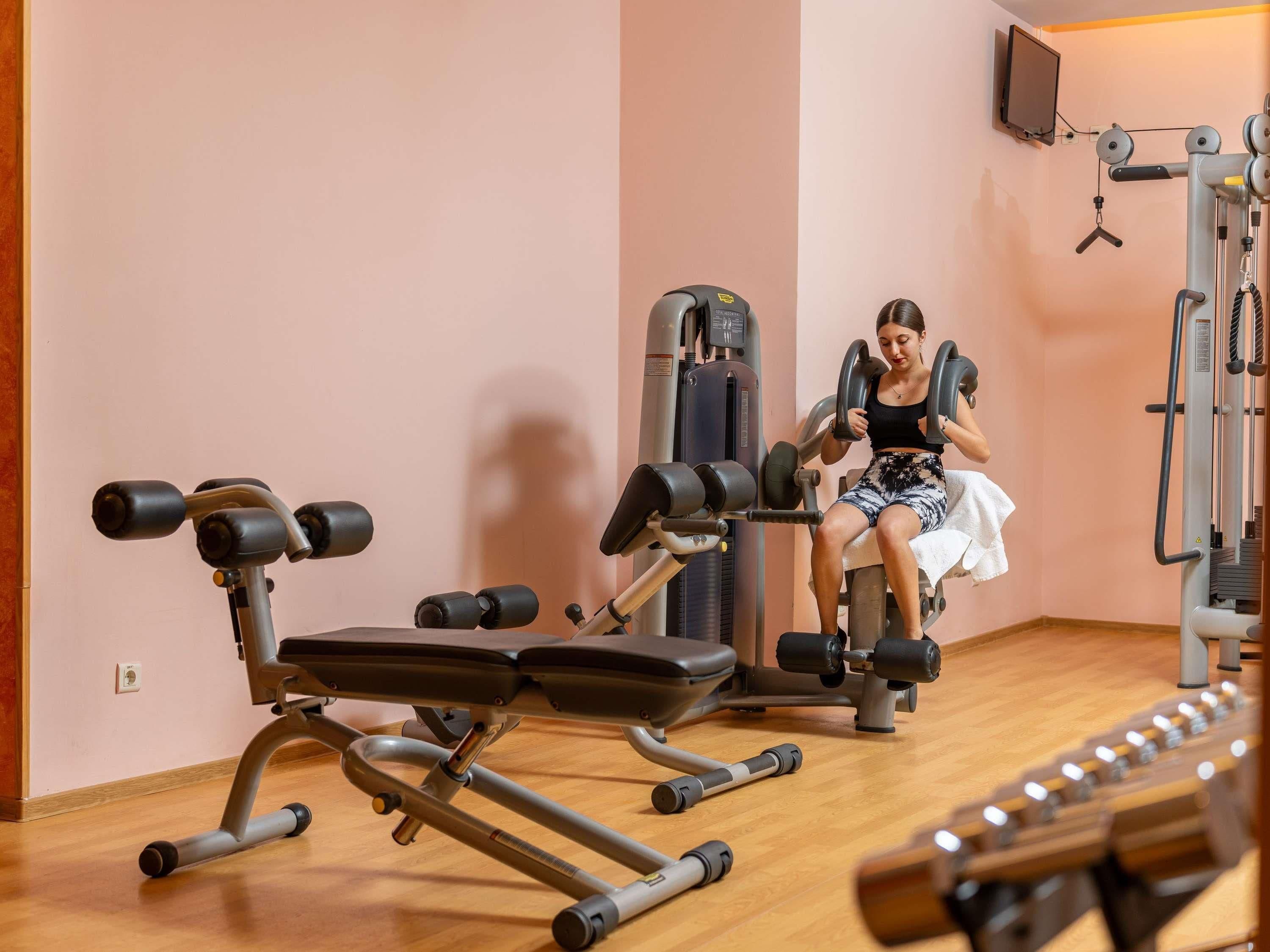 Uma academia com equipamentos de exercícios, um banco e uma televisão fixada na parede.