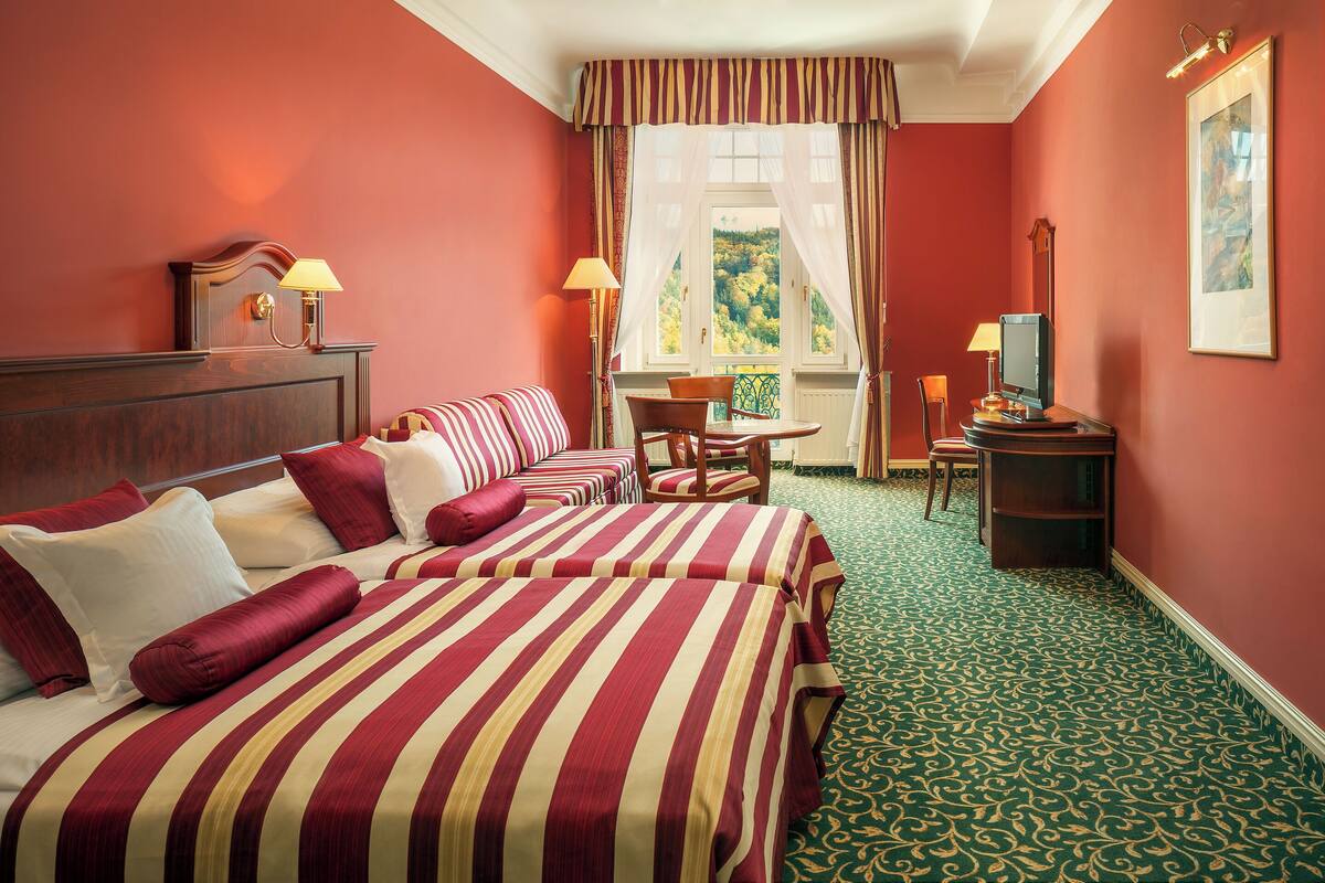 Quarto de hotel com uma cama grande, uma escrivaninha com cadeira, uma mesinha e uma janela com cortinas.