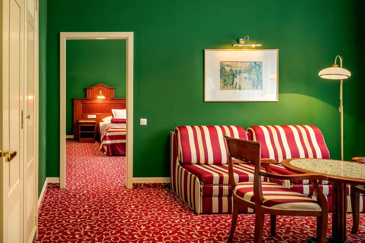 Quarto com parede verde, sofá listrado, cadeira de madeira, mesa pequena e cama com cabeceira.