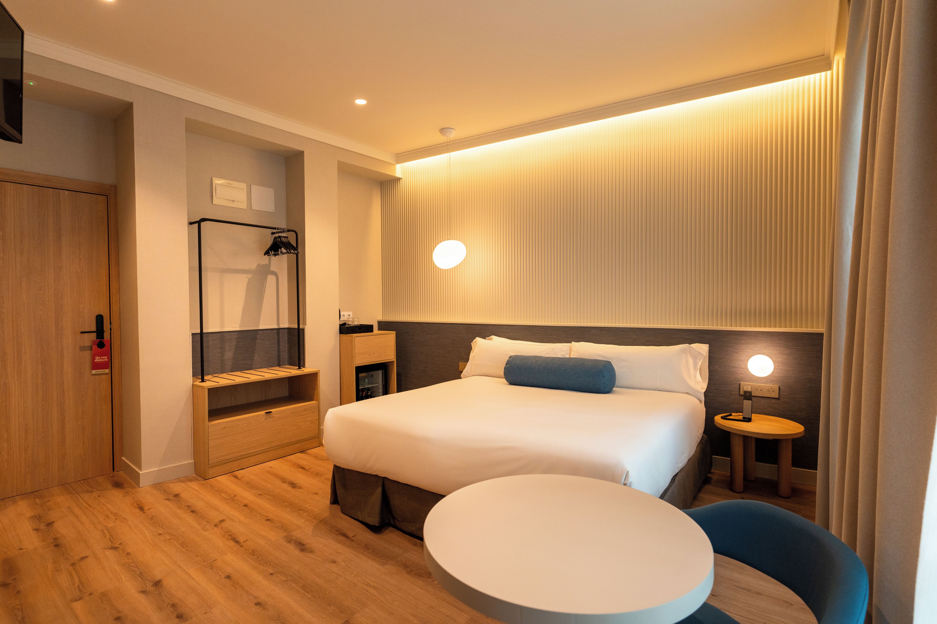 Quarto de hotel com cama, mesa de cabeceira, luminária, cadeira e guarda-roupa de madeira.