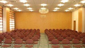Uma sala de conferência com fileiras de cadeiras vermelhas, um candelabro e uma parede de madeira com um logotipo.