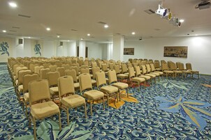 Uma sala de conferências com fileiras de cadeiras bege, carpete estampado e projetor instalado no teto.