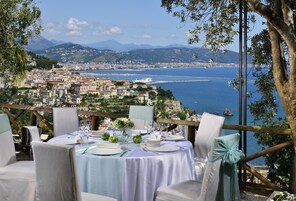 Mesa posta para refeição, com vista para uma cidade costeira e montanhas ao fundo.