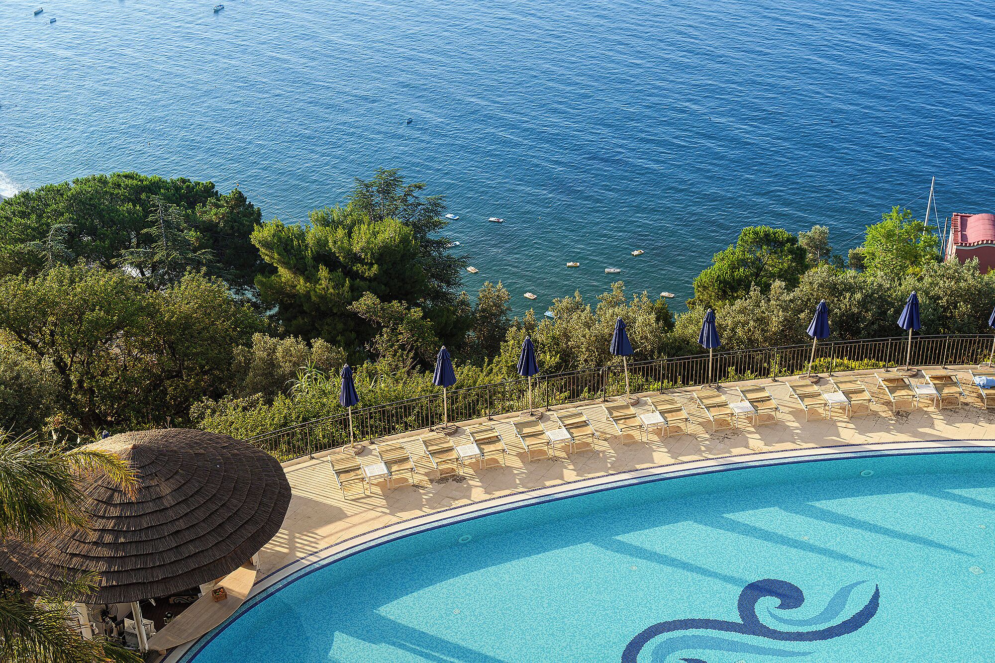 Uma piscina com um design espiral azul, rodeada por espreguiçadeiras e guarda-sóis, com vista para um corpo de água e barcos.