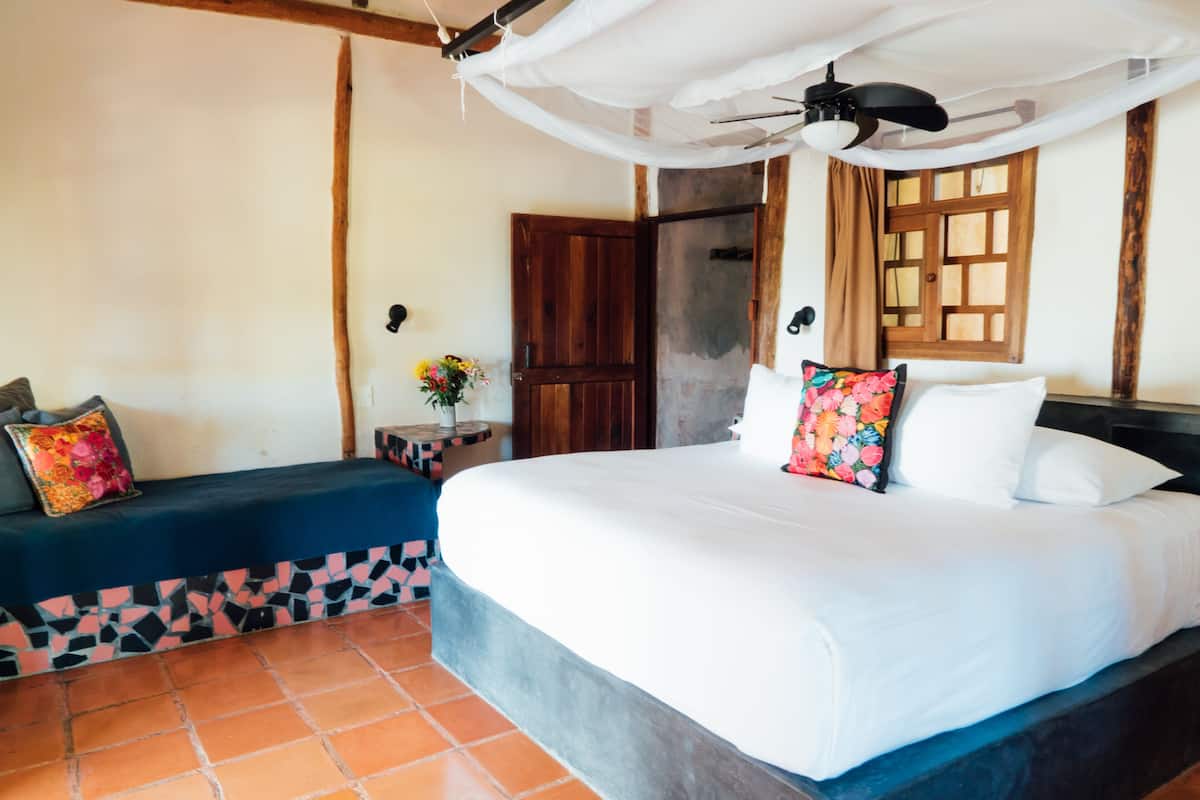 Quarto com cama, um banquinho, ventoinha no teto, janela com cortinas e porta.