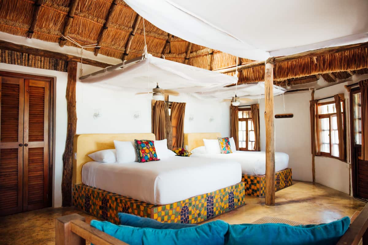 Quarto com duas camas, teto de colmo, vigas de madeira e piso quadriculado.