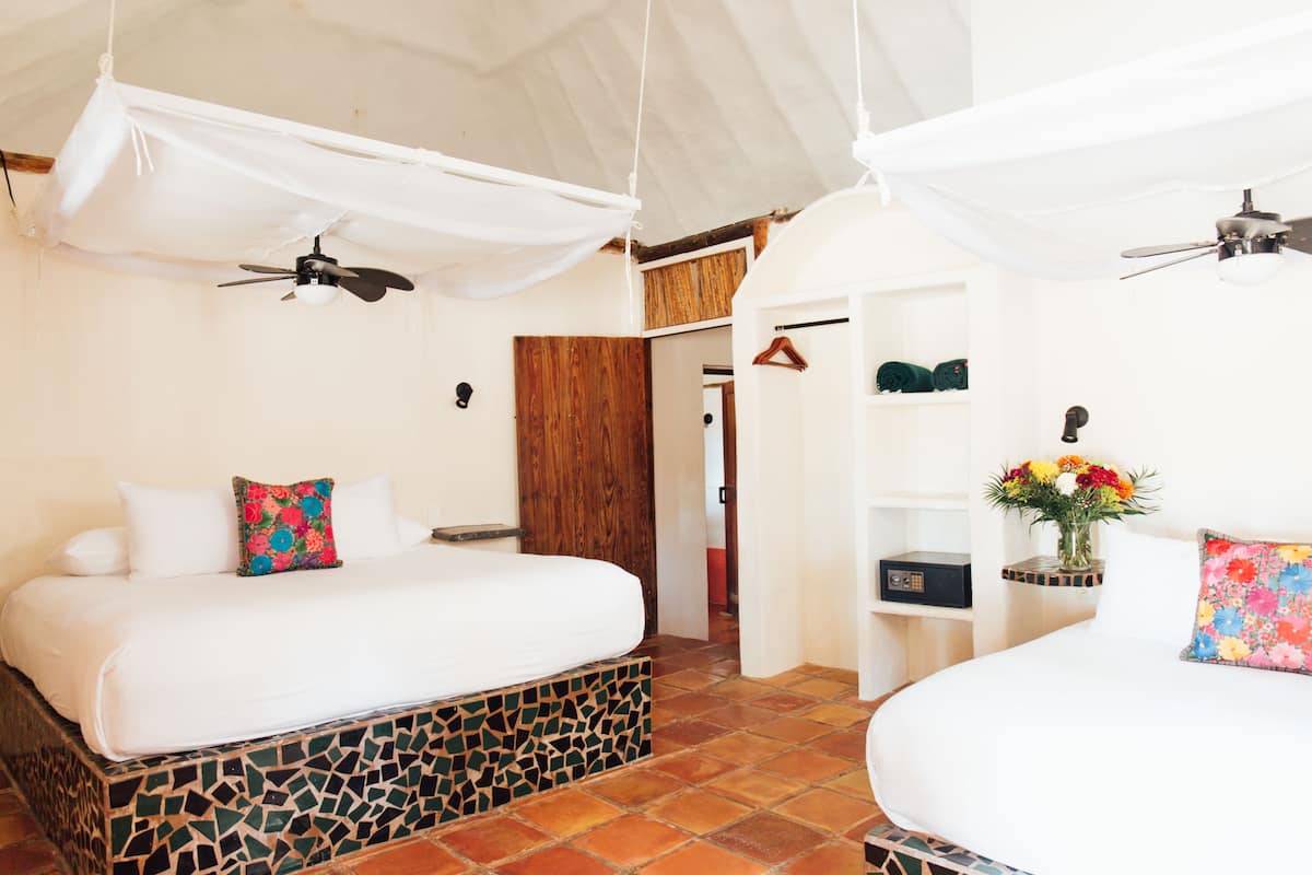 Quarto com duas camas, um ventilador de teto, porta de madeira, prateleira com um vaso de flores e piso de azulejos.