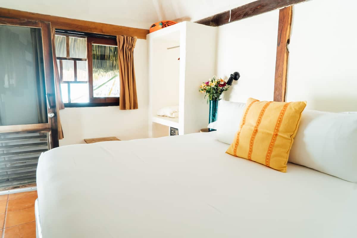 Quarto com uma cama grande, cabeceira de madeira, janela com cortinas, mesa de cabeceira e um vaso com flores.