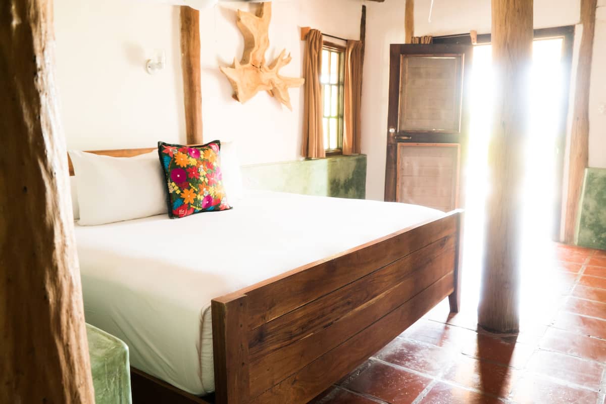 Uma cama de madeira com colchão branco e almofadas coloridas, decoração rústica na parede e uma janela com cortinas.