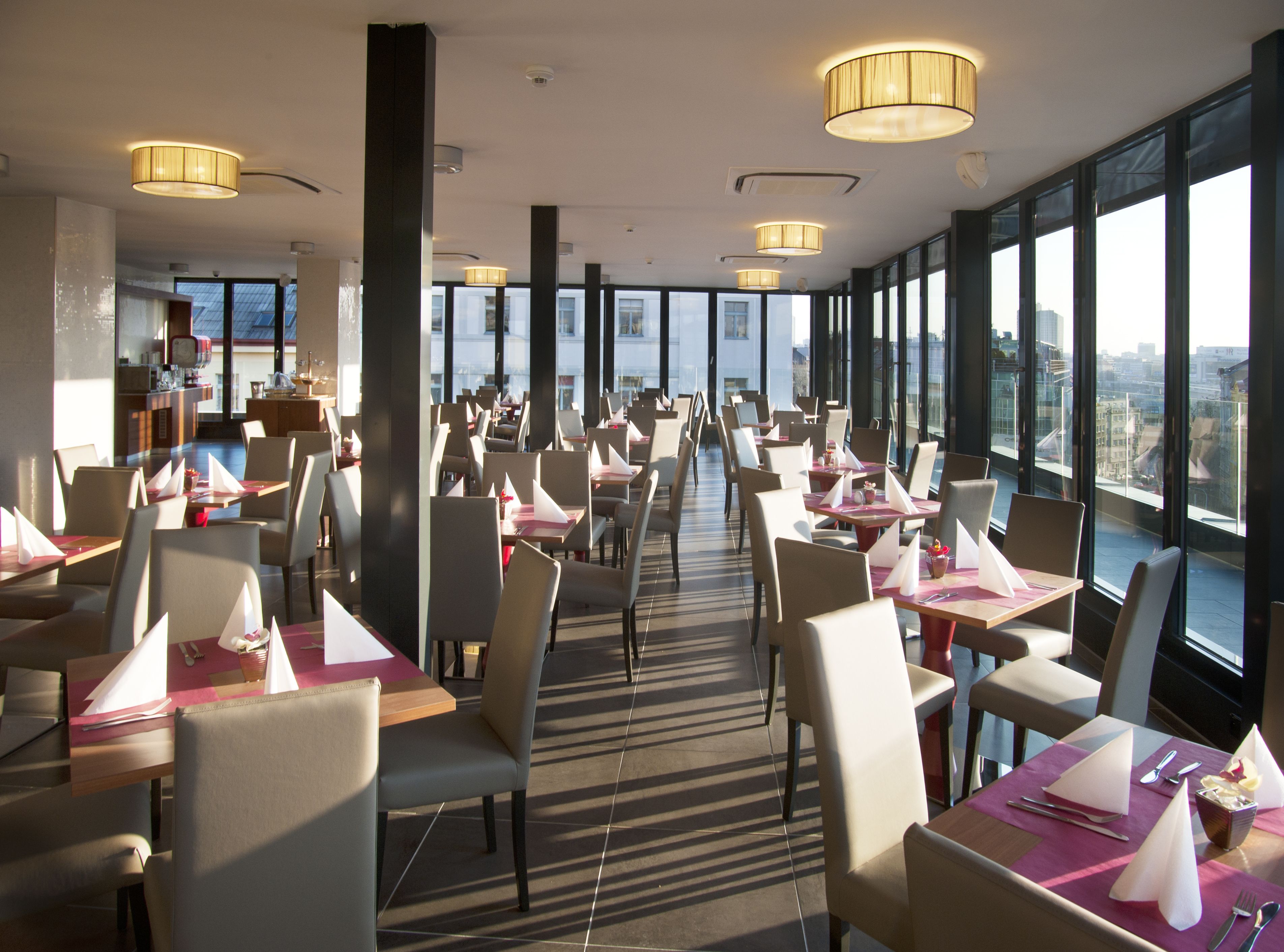 Um restaurante moderno com teto alto, grandes janelas e vista para a cidade. Mesas organizadas com guardanapos e talheres.