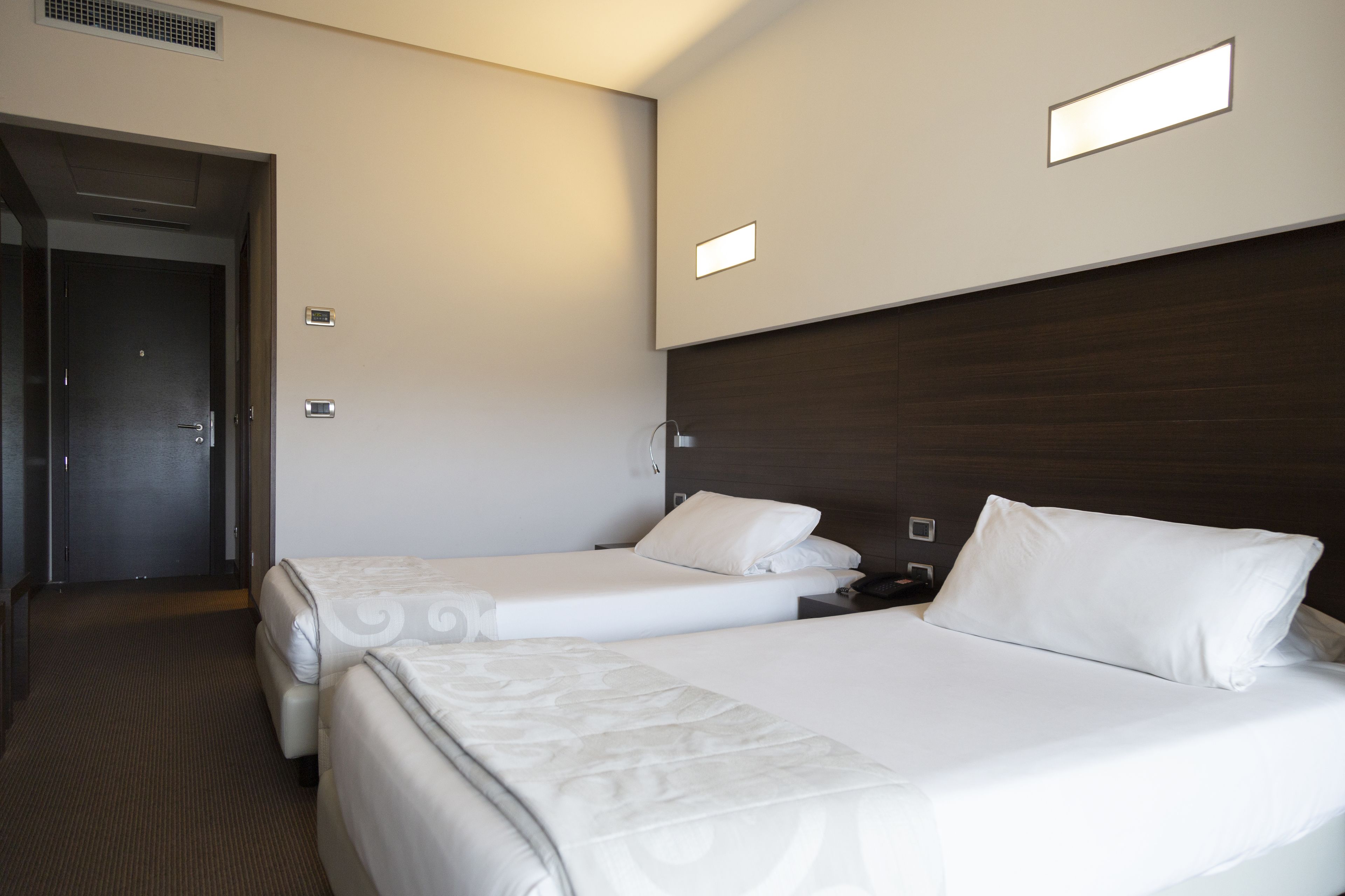 Quarto de hotel com duas camas, cabeceira em madeira escura, piso coberto por carpete e porta que dá para outro cômodo.