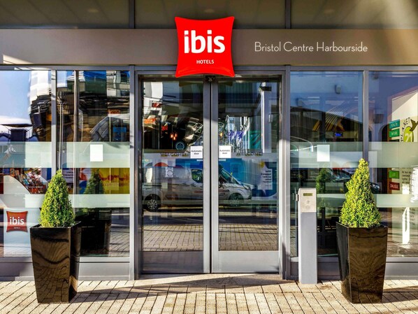 Exterior do hotel ibis com entrada de vidro, portas automáticas e duas plantas em vasos.