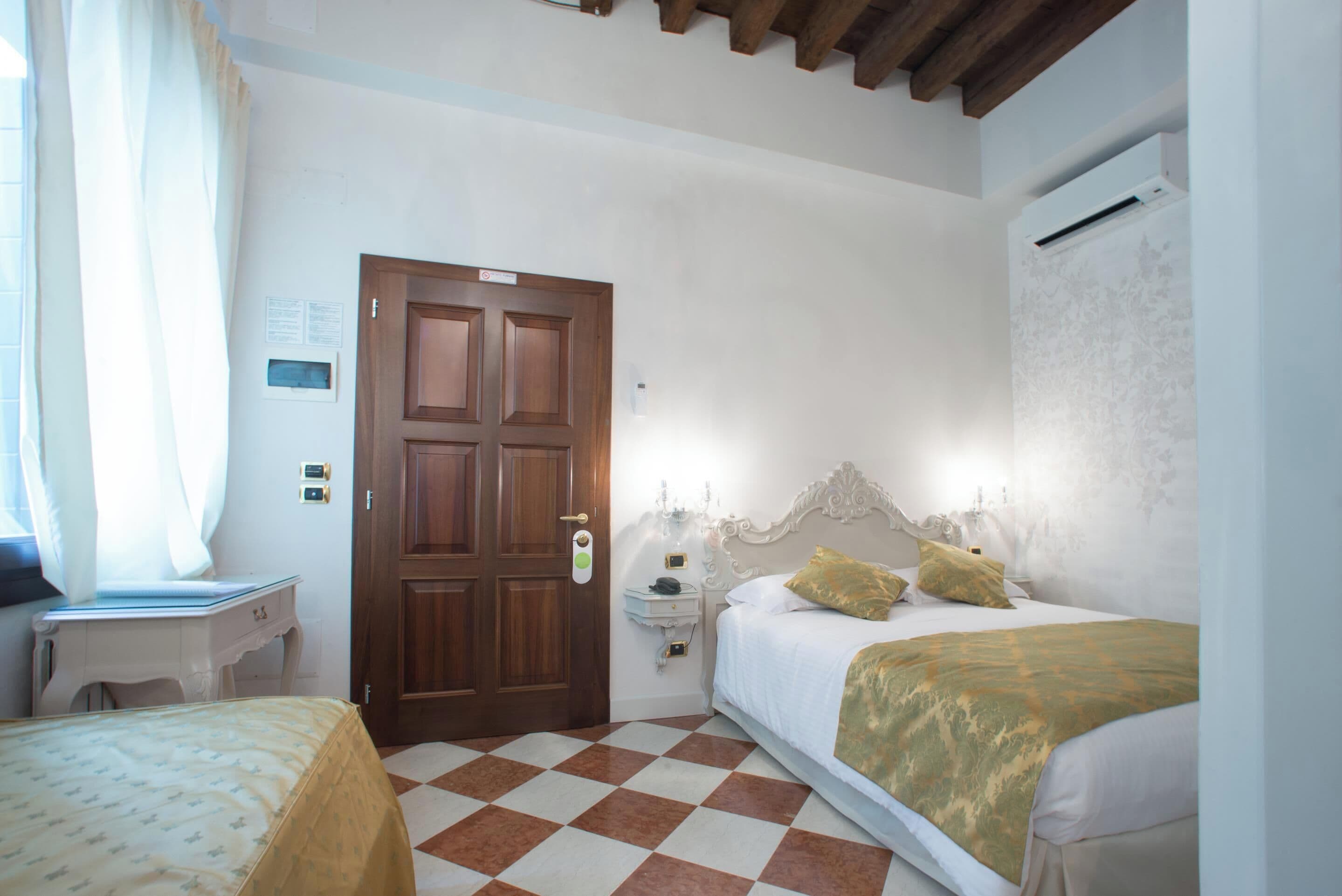 Quarto com chão xadrez, cama com colcha estampada, mesa de cabeceira com luminária e porta com fechadura.
