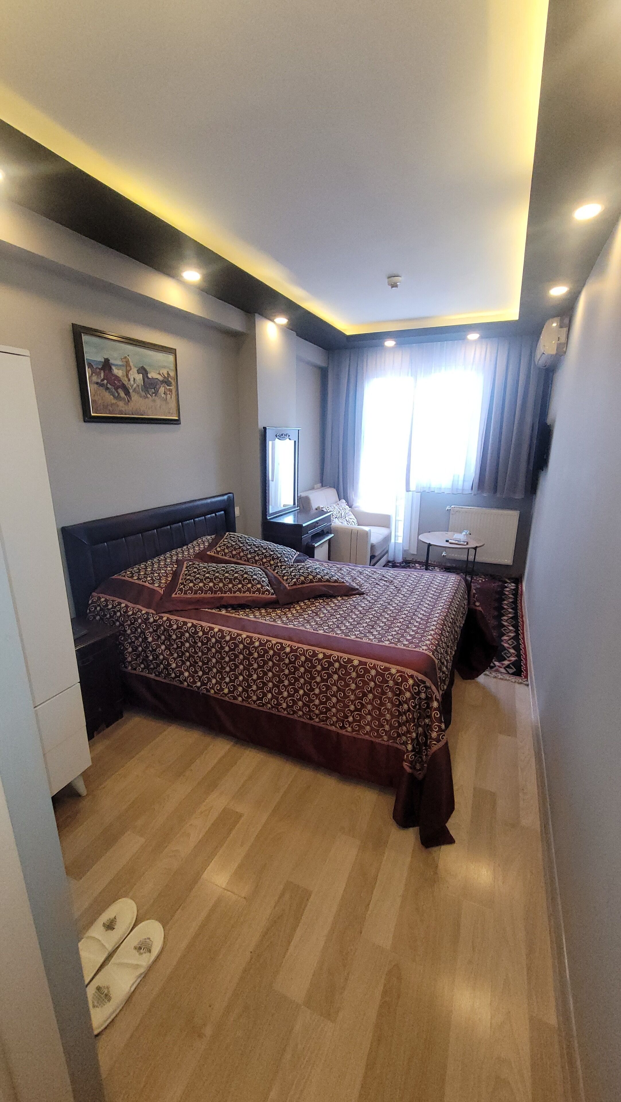 Quarto de hotel com uma cama grande, uma escrivaninha, uma cadeira, uma mesinha e uma janela com cortinas.