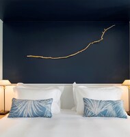 Quarto de hotel com parede azul escura, uma decoração com galhos, roupa de cama branca e duas almofadas azuis estampadas.