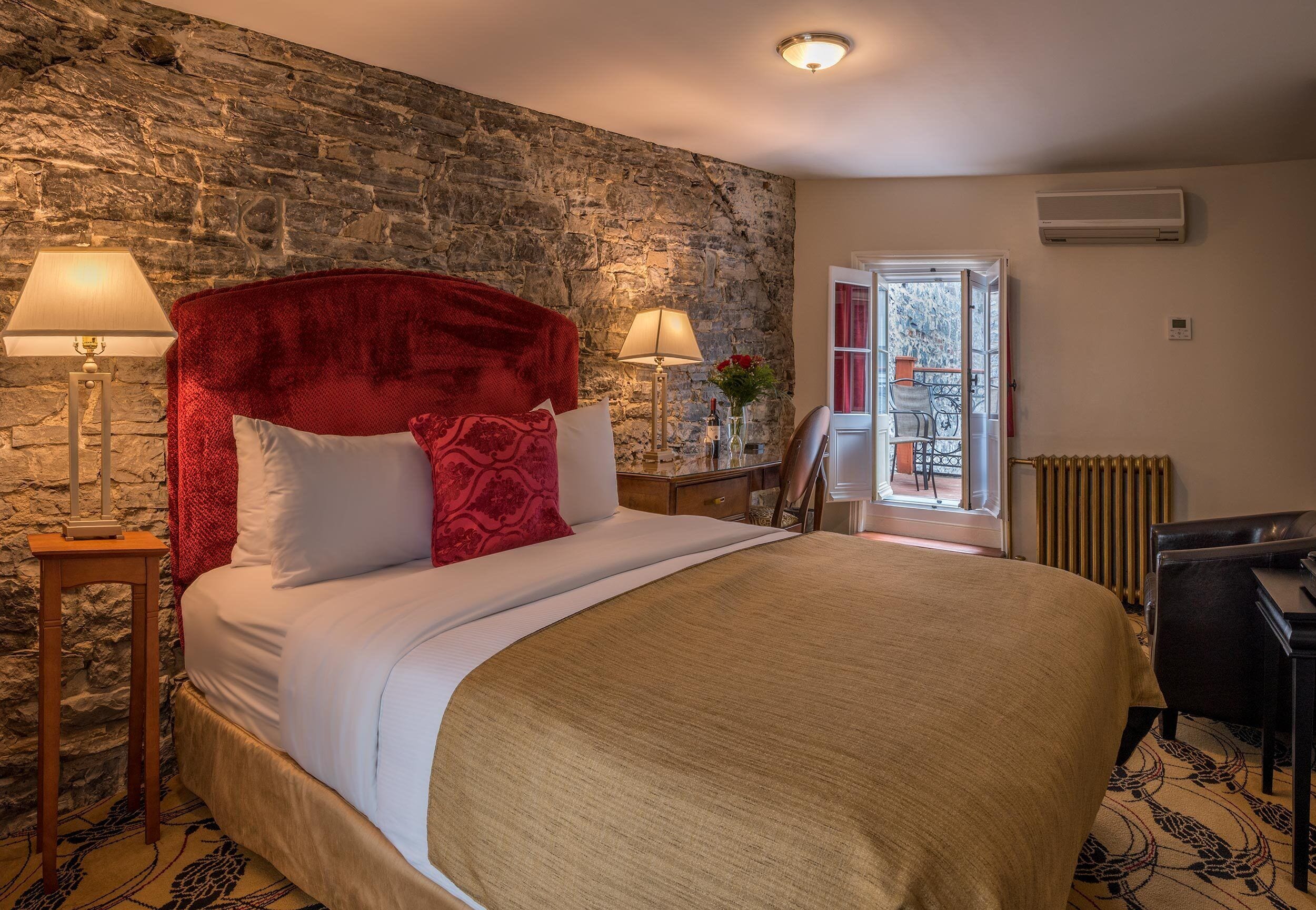 Quarto de hotel com parede de pedra, uma cama grande com roupa de cama vermelha e branca, duas luminárias de cabeceira, um radiador e vista pela janela.