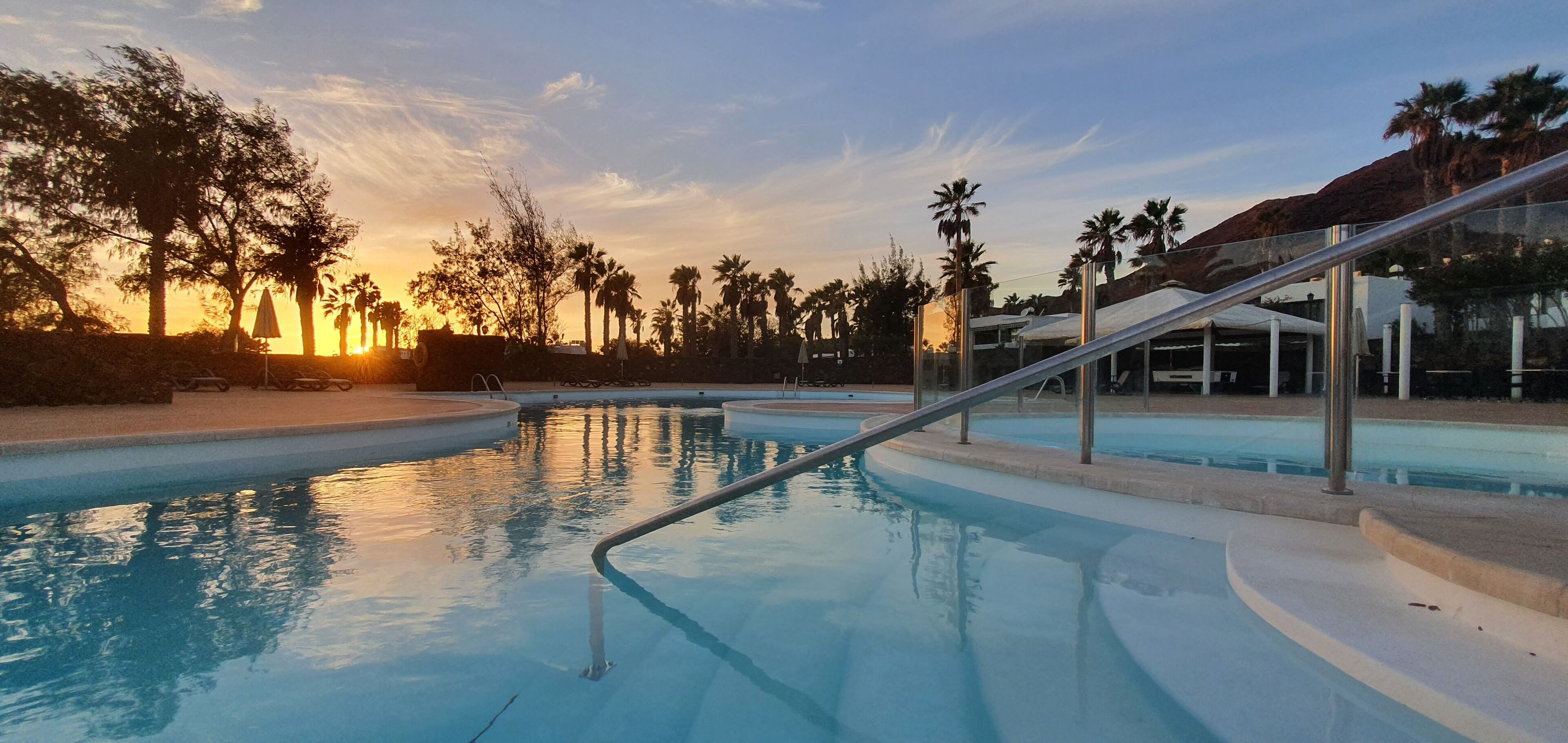 Piscina com barreira de vidro, rodeada por palmeiras e um pôr do sol ao fundo.