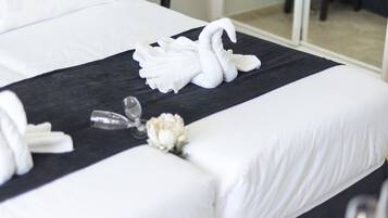 Quarto de hotel com cama bem arrumada, mesa de cabeceira com telefone, uma pequena tigela de frutas e um vaso com flores.
