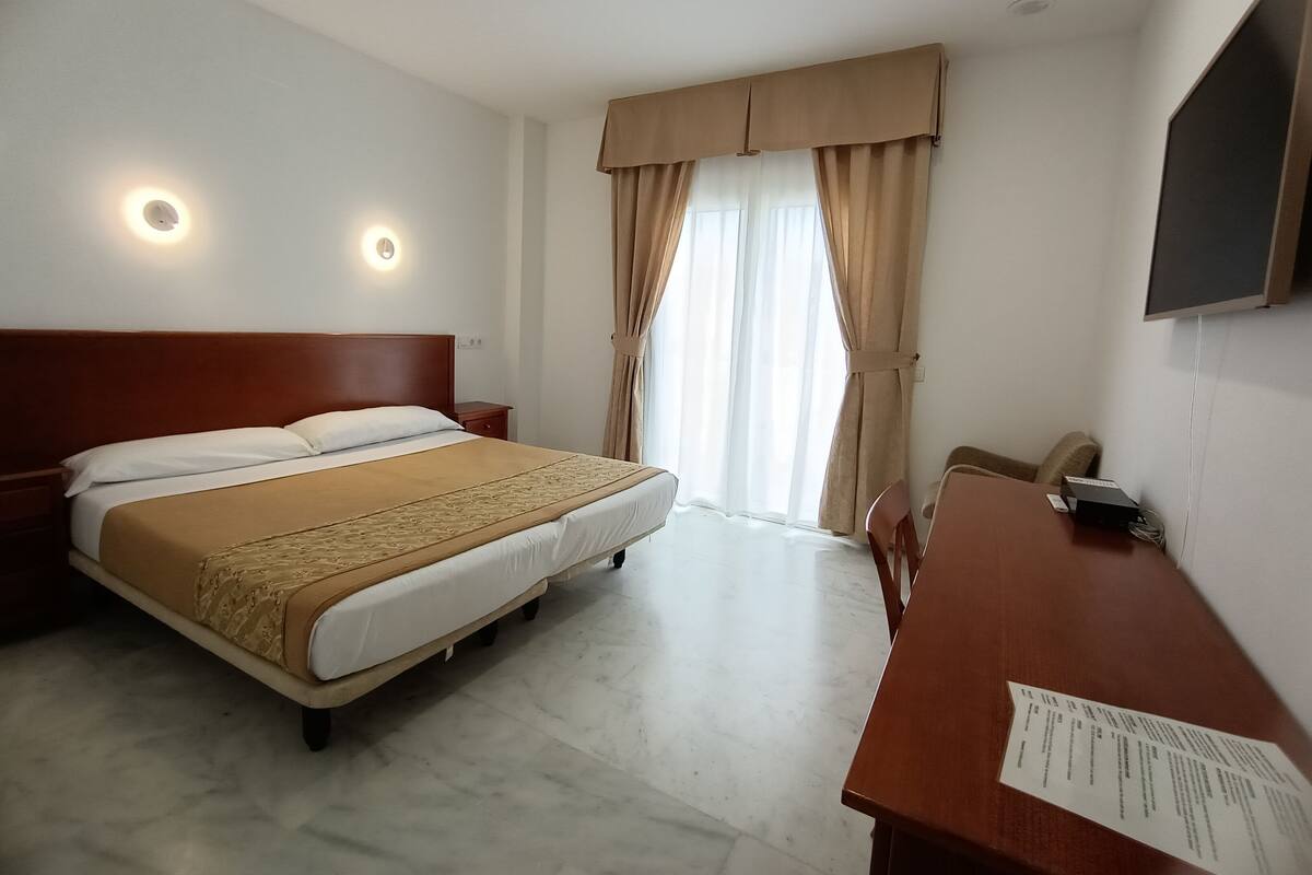 Quarto de hotel com cama, mesa de cabeceira e escrivaninha com cadeira. Possui duas luminárias fixadas na parede e uma janela com cortinas.