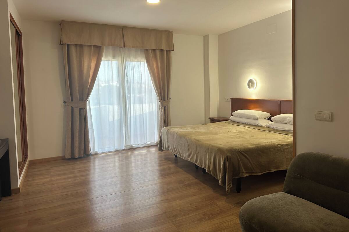 Quarto de hotel com cama, mesa de cabeceira, cadeira, janela com cortinas e luminária no teto.
