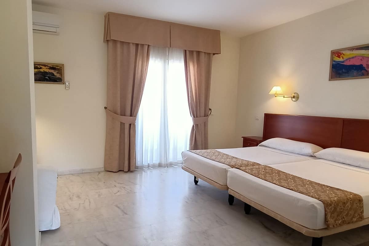 Quarto de hotel com cama de casal, mesa de cabeceira, escrivaninha com cadeira, janela com cortinas e um quadro na parede.