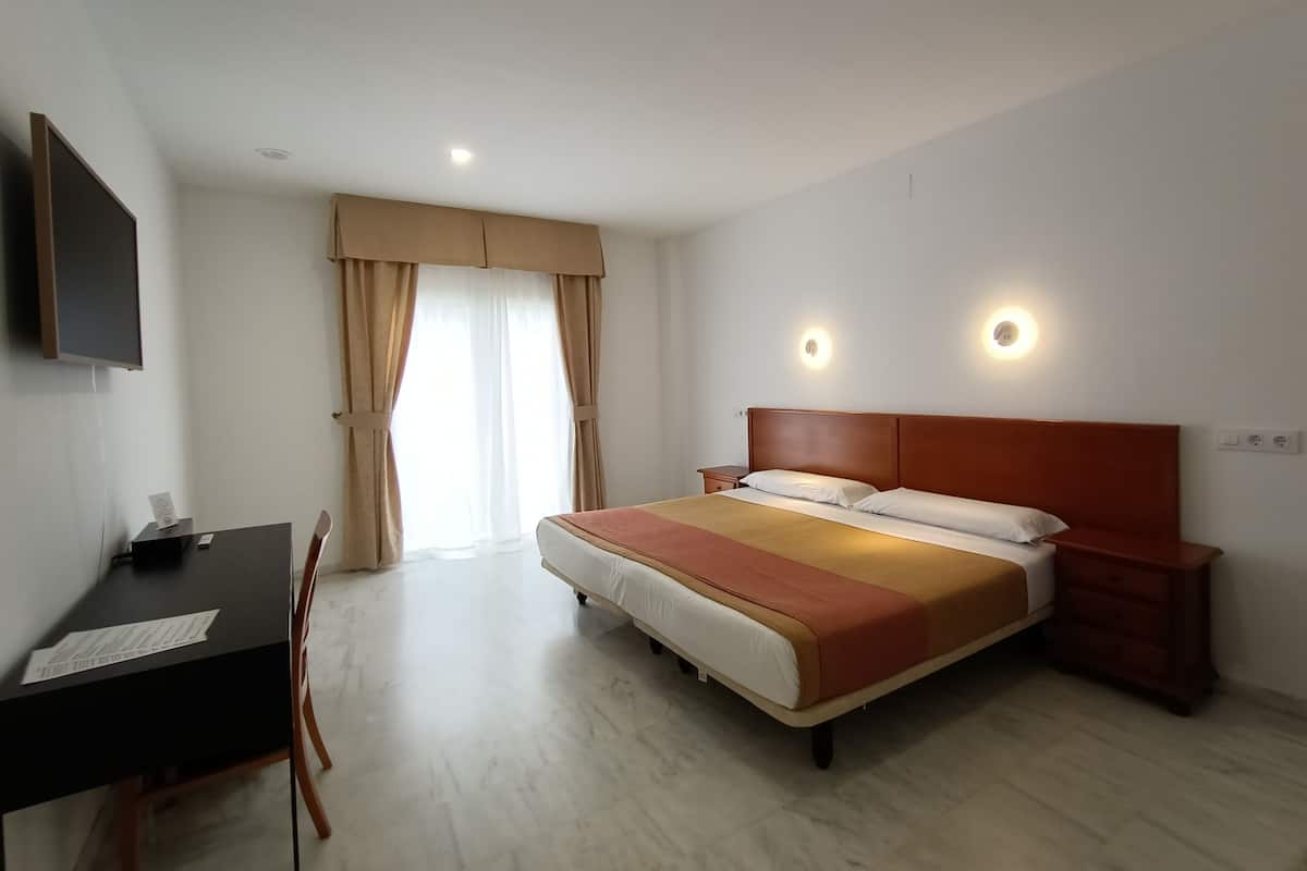 Quarto de hotel com cama, mesa de cabeceira, escrivaninha, cadeira e televisão fixada na parede.