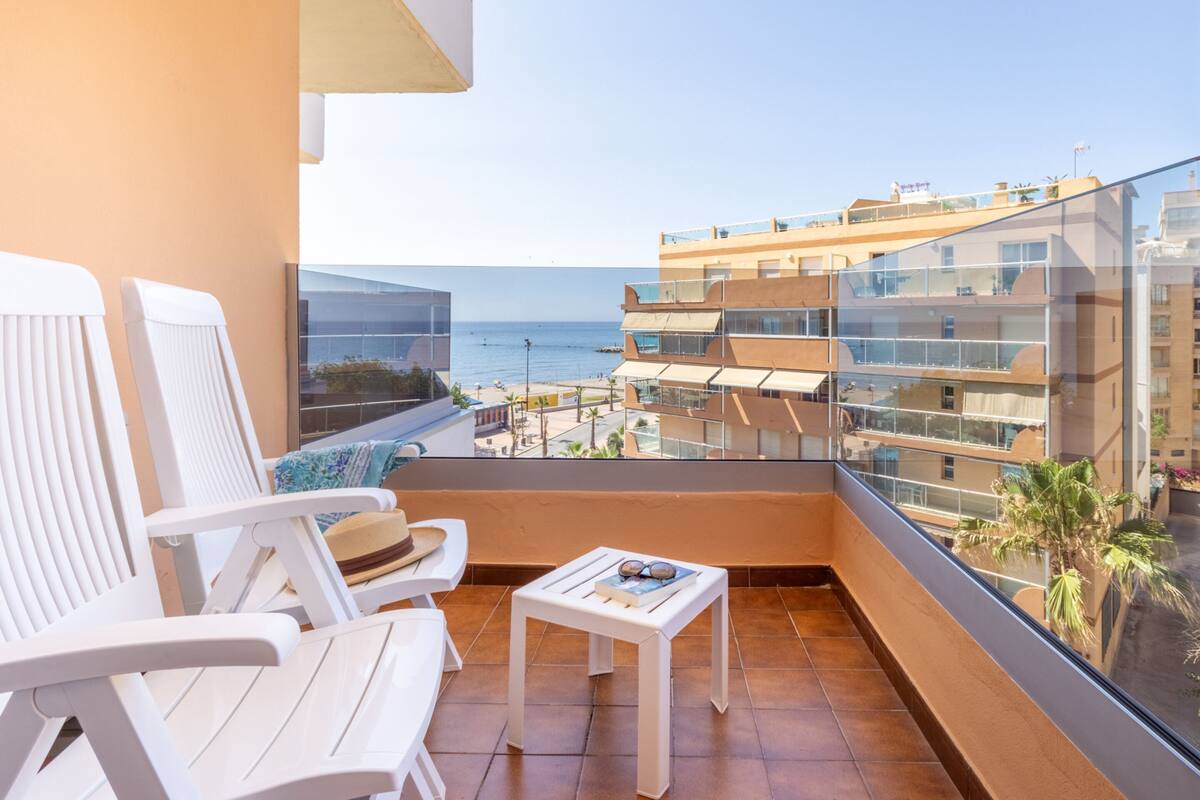 Varanda com cadeiras brancas e uma mesa pequena, com vista para a praia e outros edifícios.