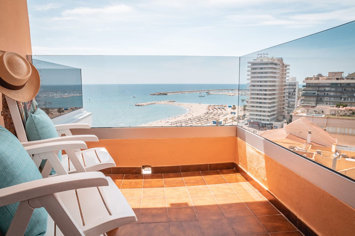 Uma varanda com cadeiras brancas, um chapéu e vista para a praia e edifícios.