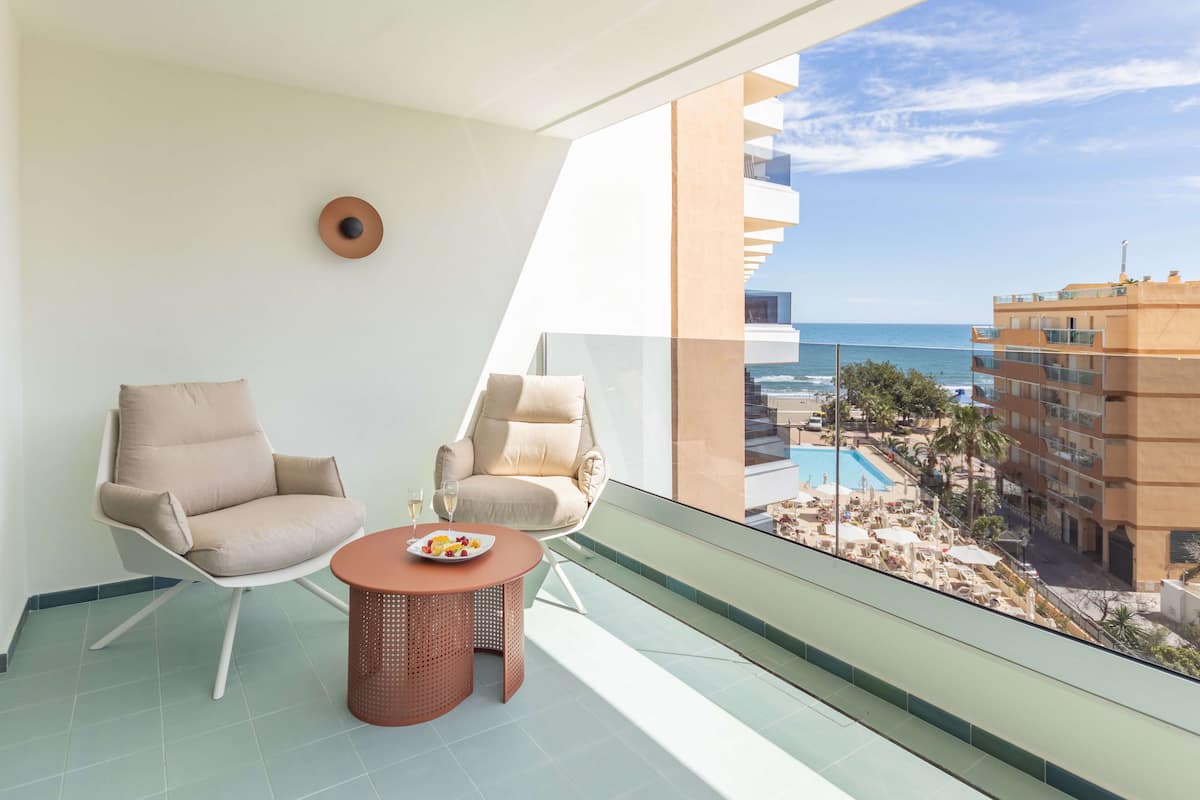 Varanda com duas cadeiras e uma mesa pequena, com vista para uma piscina e prédios.