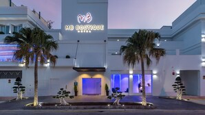 Um hotel moderno com uma placa MB Boutique em destaque, entrada bem iluminada e paisagismo frontal com palmeiras.