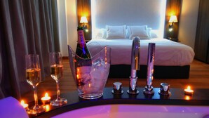 Quarto de hotel com cama, luminárias de cabeceira, minibar e banheira com velas.