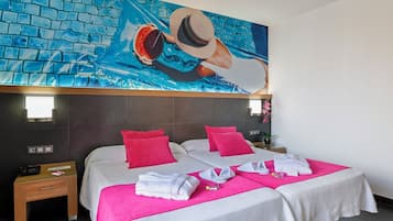 Quarto de hotel com duas camas, cada uma com uma almofada rosa, uma mesa de cabeceira de vidro com um telefone e uma toalha rosa vibrante.