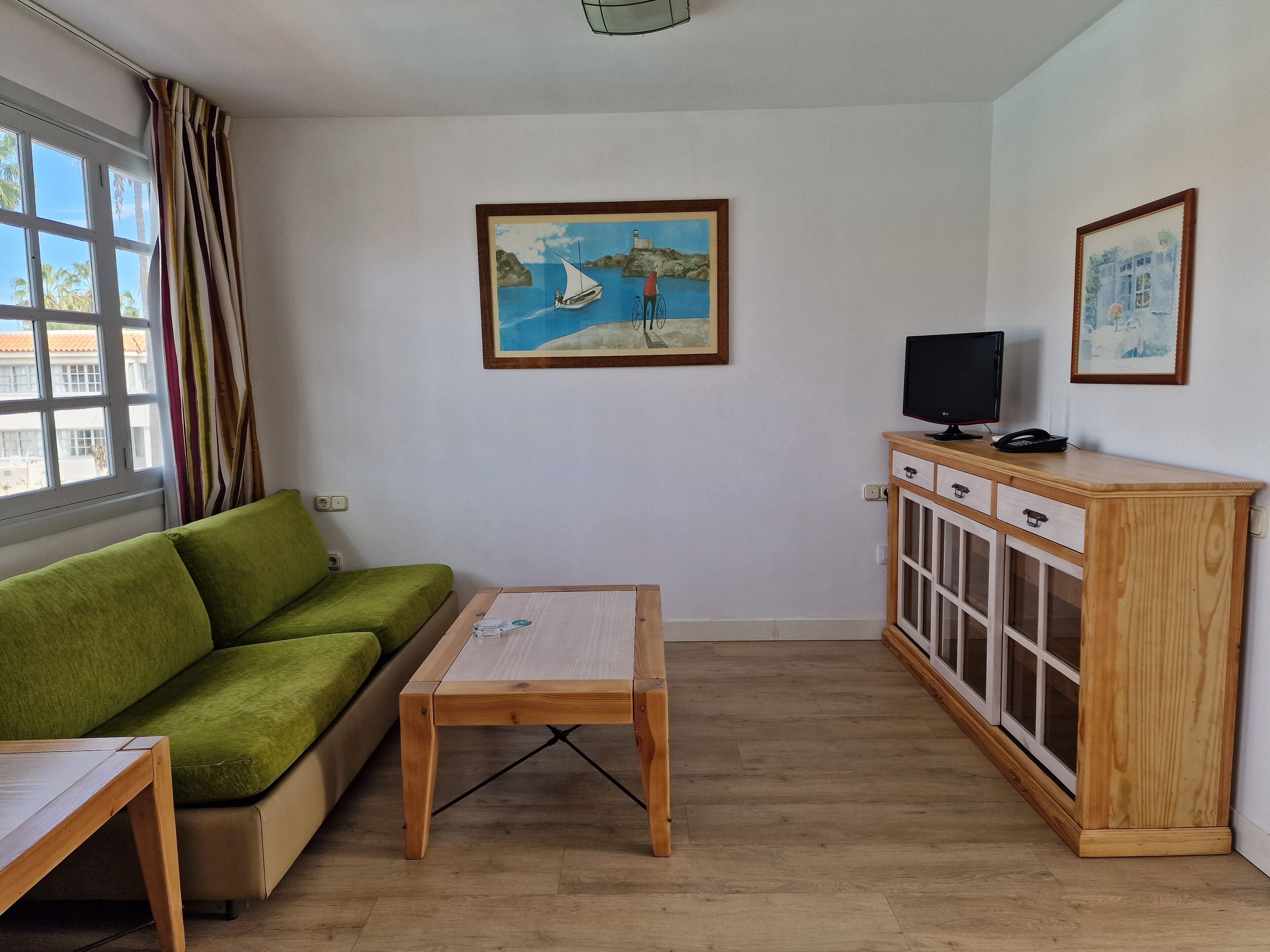 Um quarto com um sofá verde, uma mesa de madeira, um armário com gavetas, uma televisão e duas pinturas emolduradas na parede.
