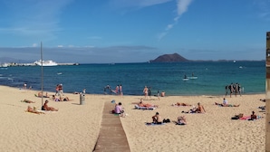 Uma praia com pessoas se banhando e tomando sol, um píer com barcos, e uma montanha ao fundo.