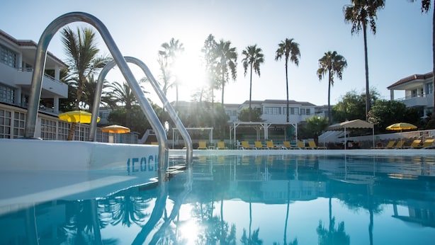 Piscina com água cristalina refletindo palmeiras e um edifício ao fundo.